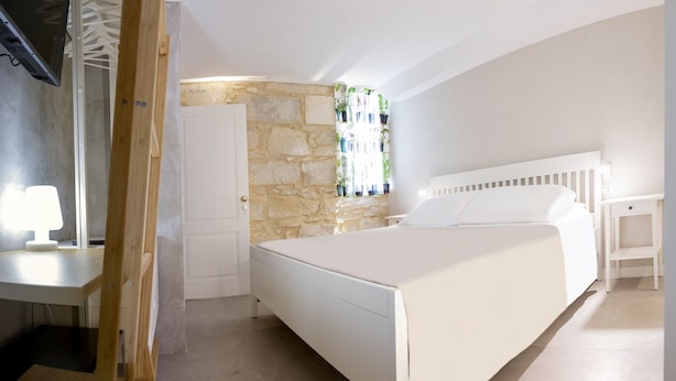 Quarto com parede de pedra, cama com roupa de cama branca, mesa de cabeceira e janela com plantas em vasos.