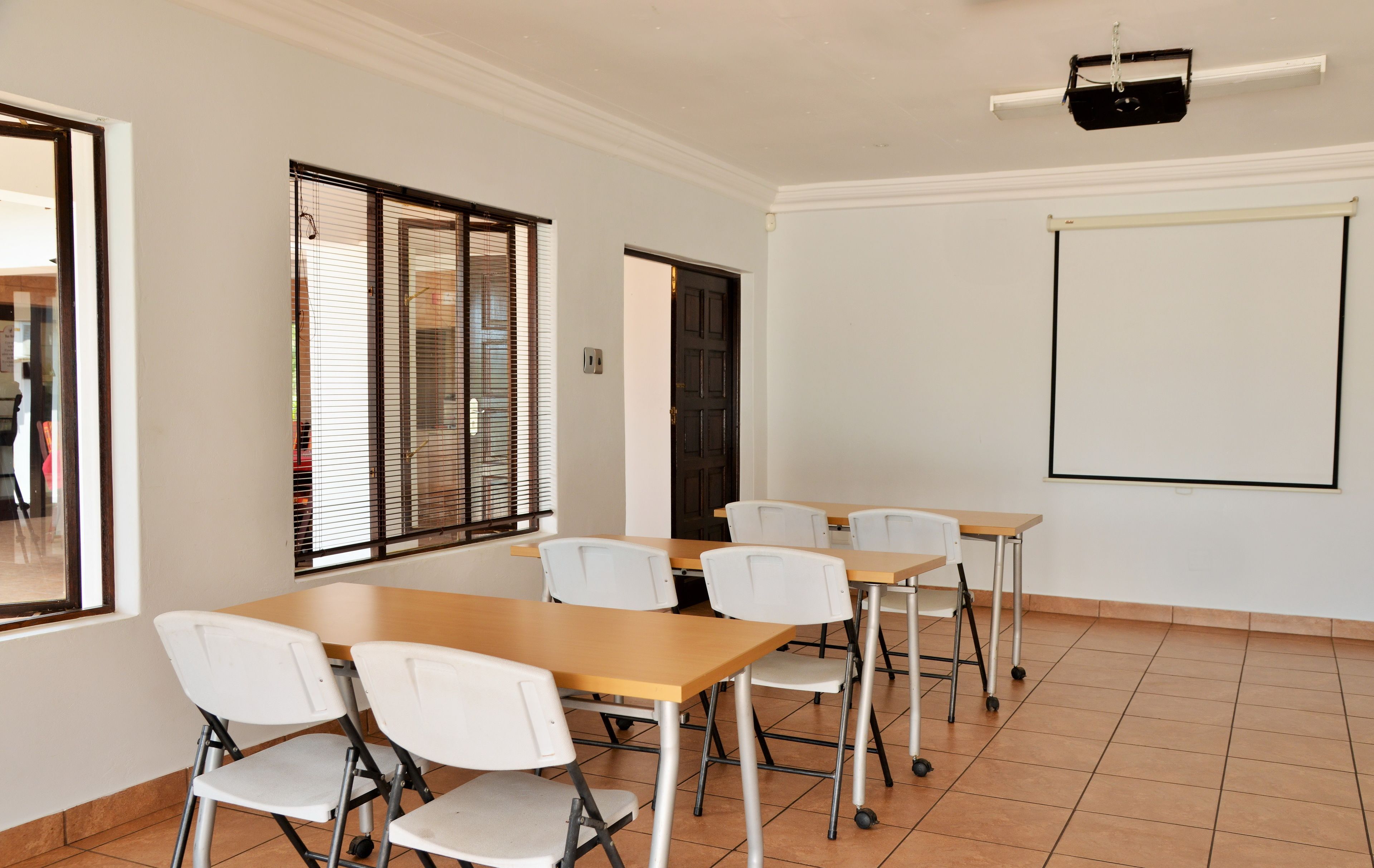 Uma sala de aula com cadeiras brancas, uma mesa de madeira, um projetor e uma lousa.