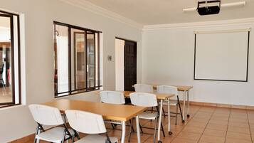 Uma sala de aula com cadeiras brancas, uma mesa de madeira, um projetor e uma lousa.