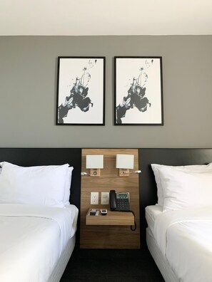 Quarto de hotel com duas camas, cabeceira de madeira com telefone e luminárias, e duas obras de arte abstratas emolduradas na parede.