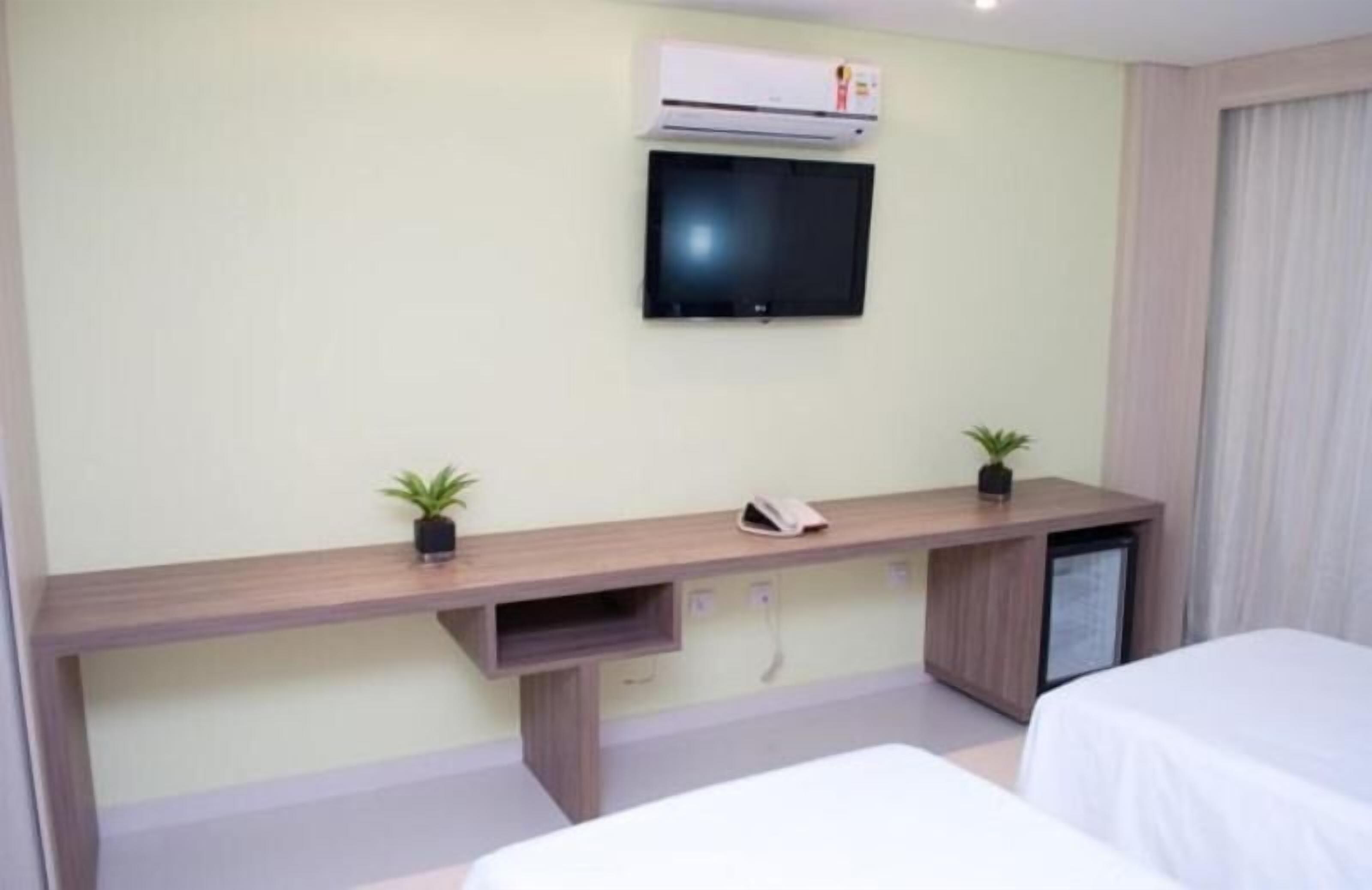 Quarto de hotel com cama, escrivaninha com telefone, uma planta pequena e ar condicionado.