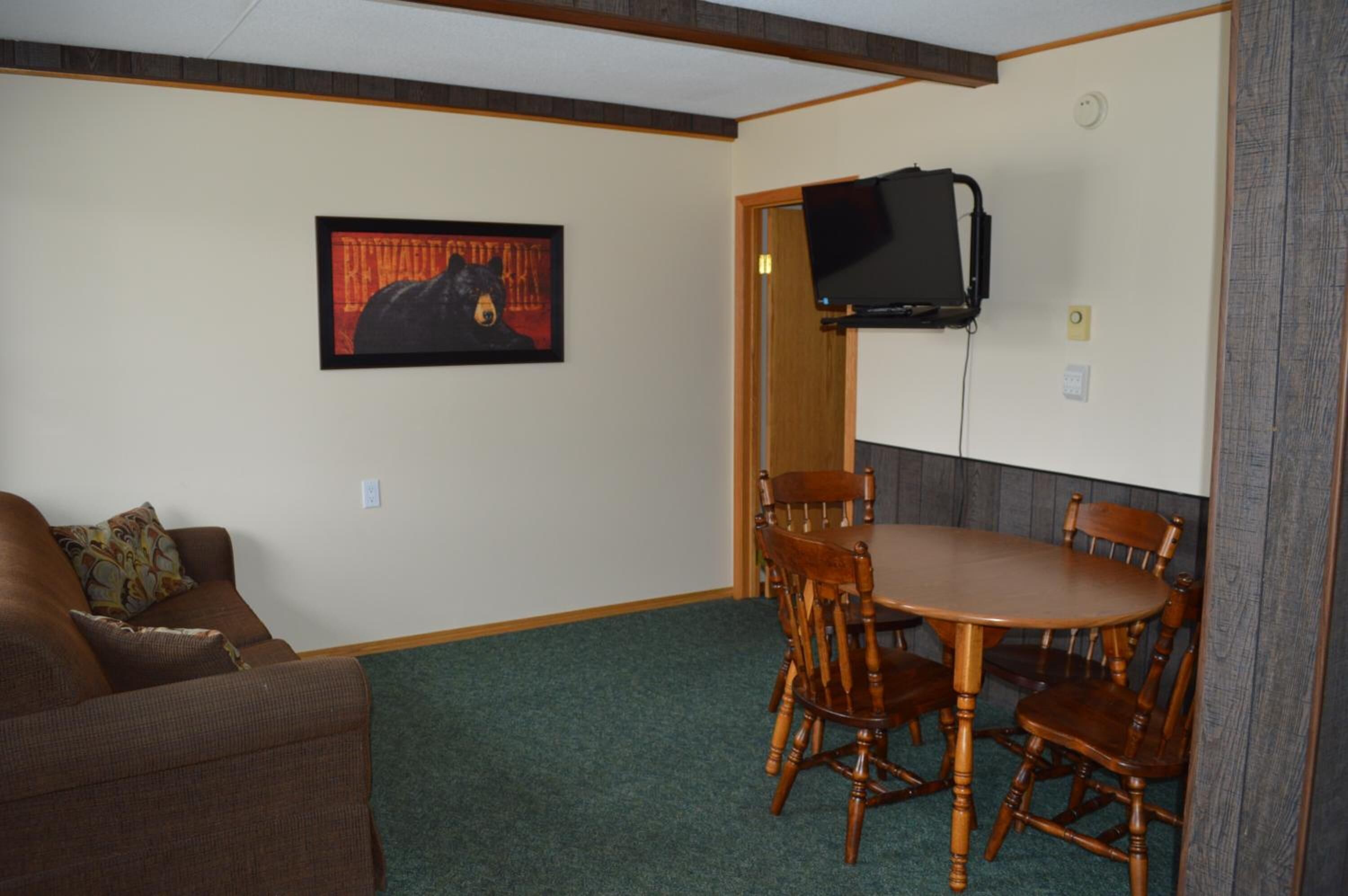 Um quarto com um sofá marrom, uma mesa de jantar de madeira com quatro cadeiras, uma televisão fixada na parede e um quadro emoldurado.