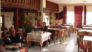 Restaurante com mesas e cadeiras de madeira, teto alto de madeira e vista para o exterior por grandes janelas.