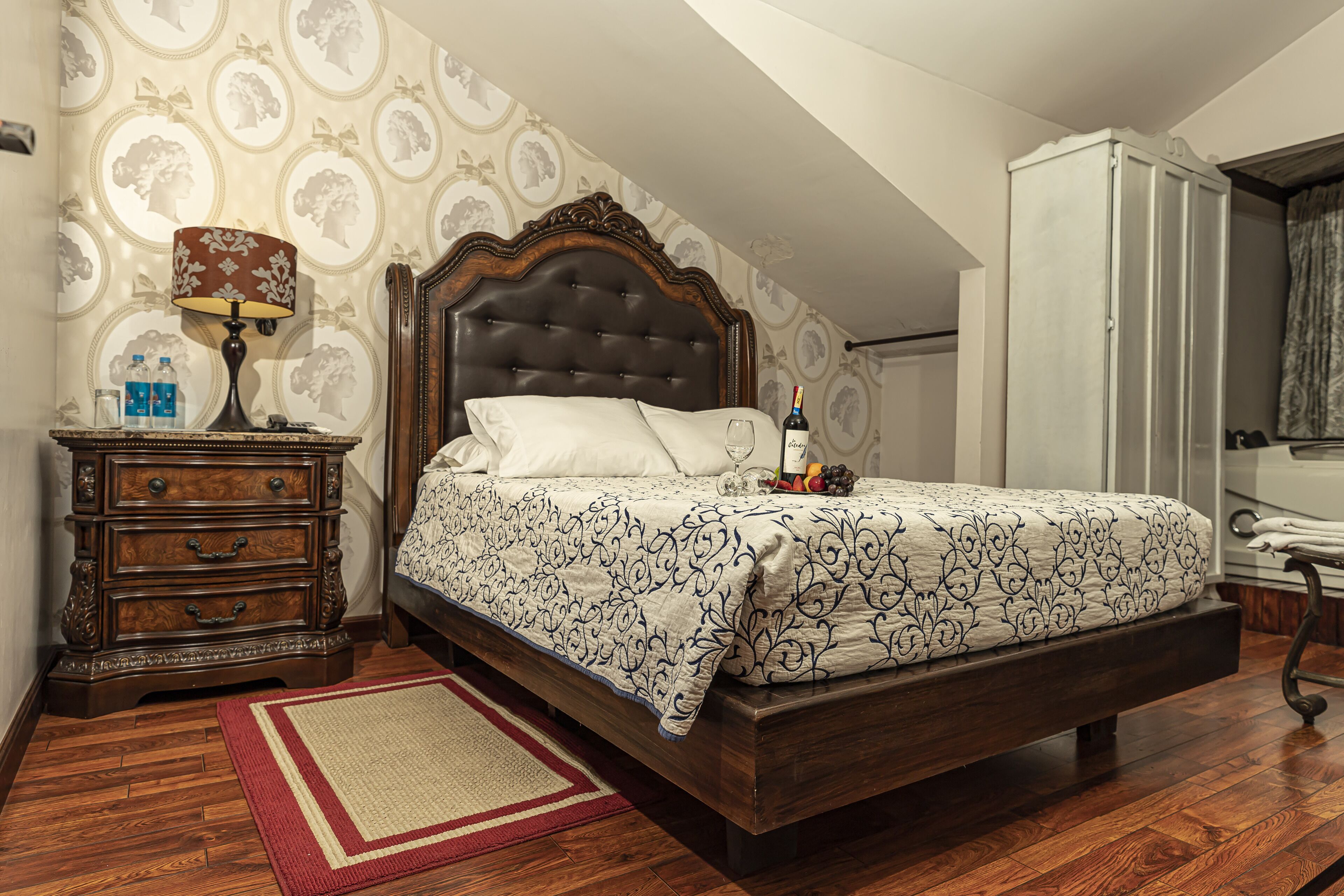 Quarto com uma cama grande, uma mesinha de cabeceira com abajur, cômoda e papel de parede estampado.