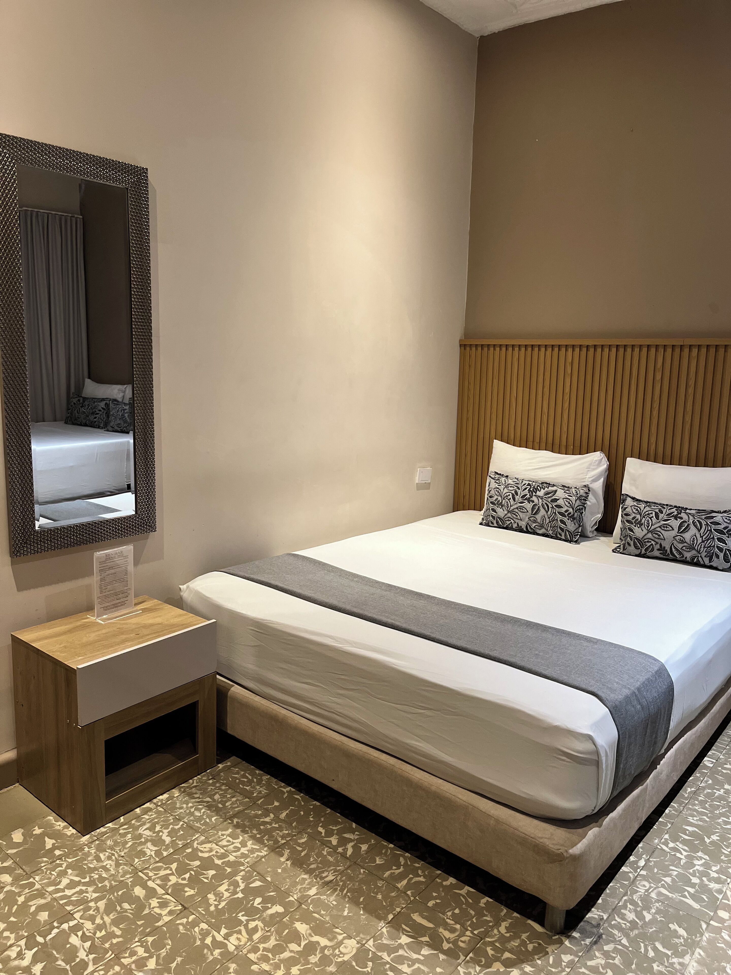 Quarto de hotel com uma cama grande, uma mesa de cabeceira, um espelho e um painel de cabeceira de madeira.