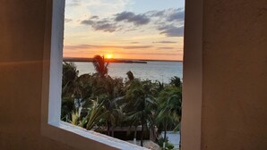 Vista do pôr do sol pela janela, com água e palmeiras.