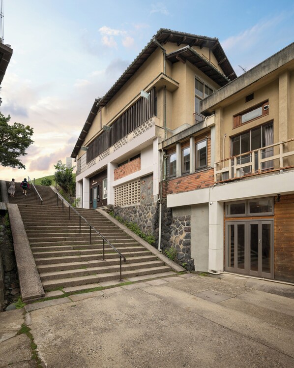 Um edifício com escadaria que dá para uma varanda no segundo andar, uma parede de pedra e uma porta de madeira.