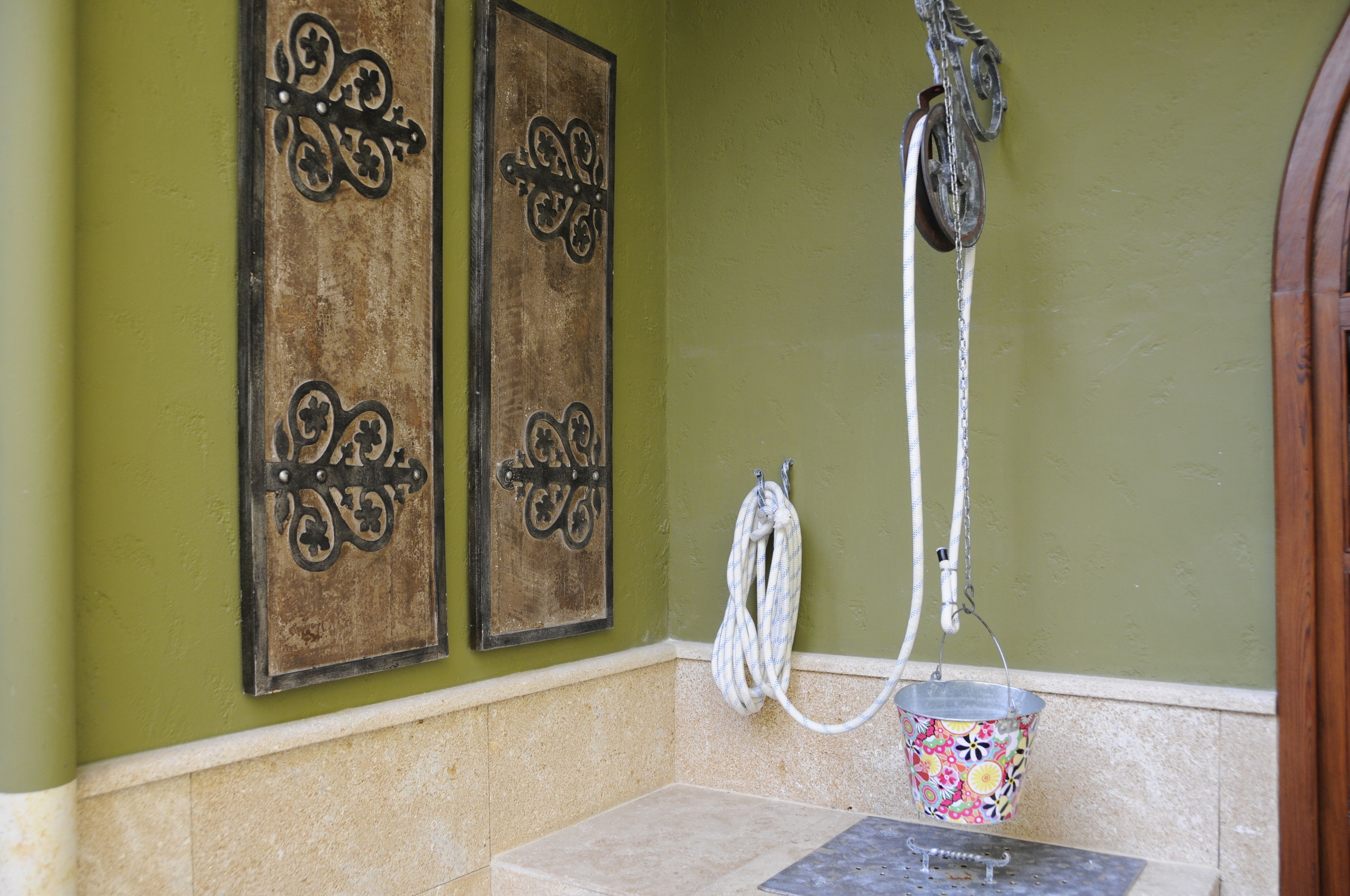 Casa de banho com parede verde, duas portas metálicas decorativas, uma mangueira branca e um balde colorido.