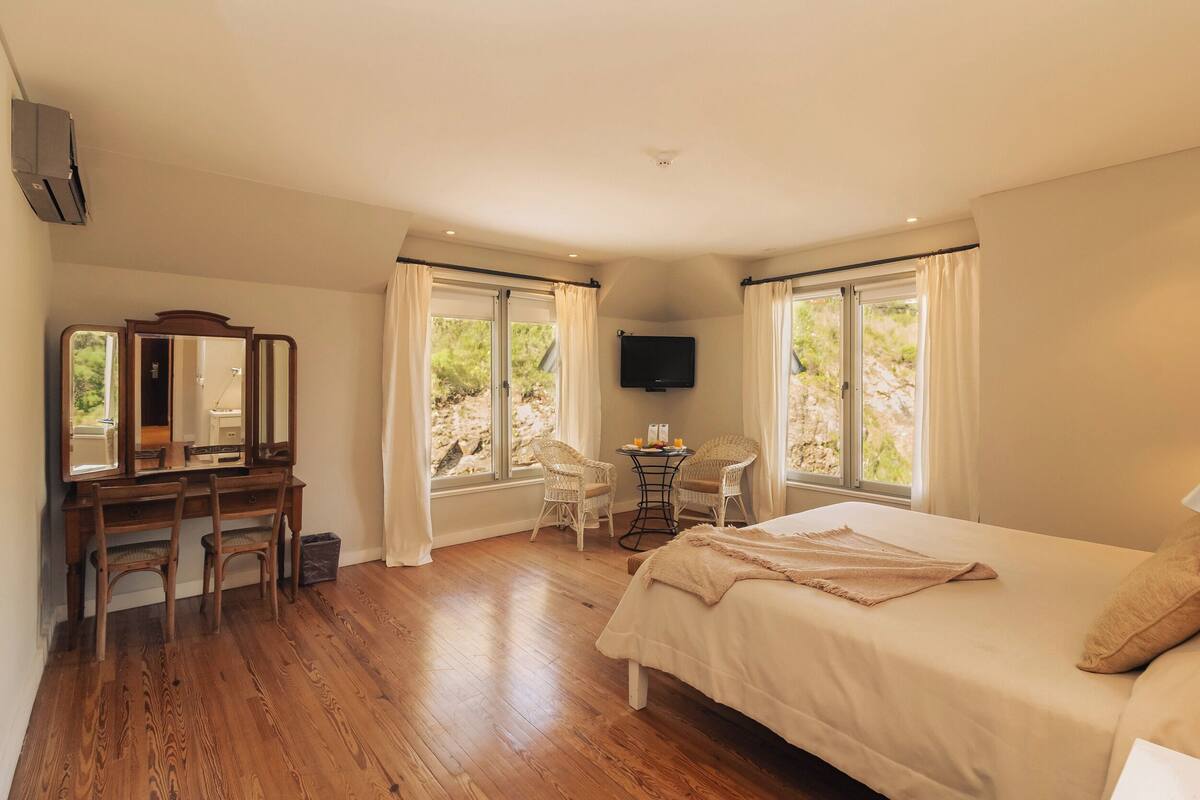 Quarto com cama, escrivaninha, cadeiras, televisão e janelas amplas.