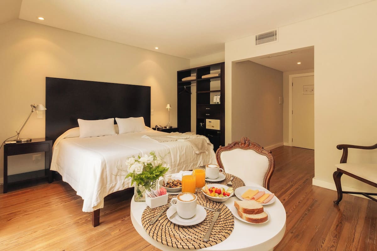 Quarto de hotel com cama bem arrumada, mesa de café da manhã com tigela de frutas, suco e café, e uma cadeira.