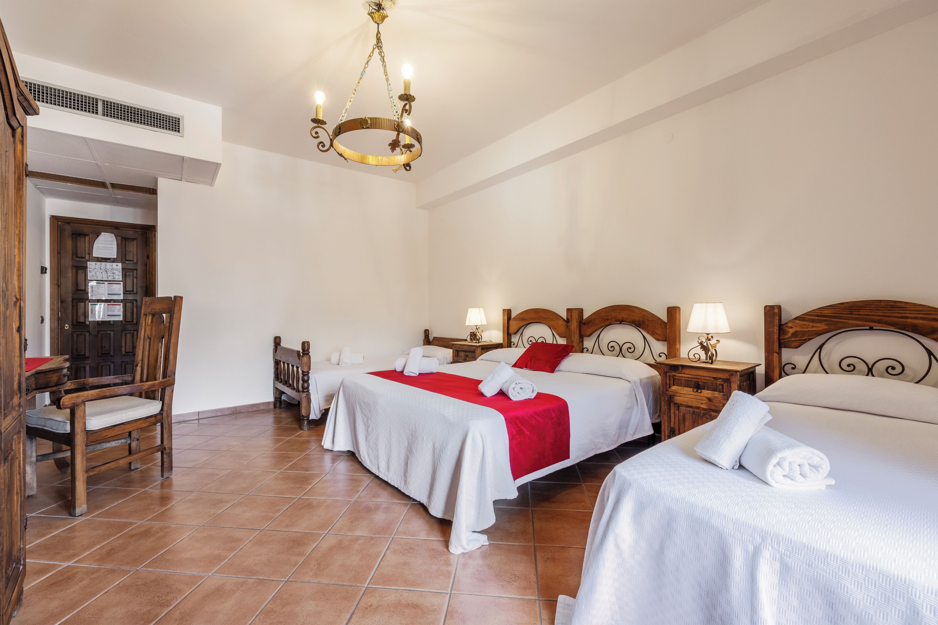 Quarto de hotel com duas camas, uma cadeira de madeira, um criado-mudo de madeira e um candelabro.