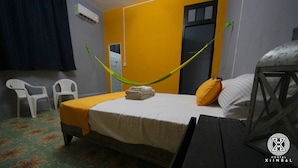 Um quarto com uma cama, uma rede, duas cadeiras de plástico brancas, uma porta e um aparelho de ar condicionado.