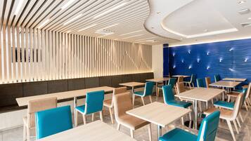 Interior de restaurante moderno com tetos de ripas de madeira, cadeiras estofadas em azul e mesas de cor clara.