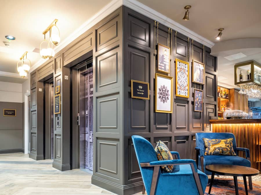 Um saguão de hotel com uma escadaria imponente, área de bar e espaço de estar com cadeiras estofadas em azul.