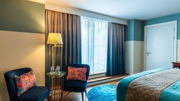 Quarto de hotel com uma cama, duas poltronas, uma mesinha de centro com abajur e uma janela grande com cortinas.