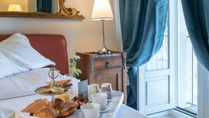 Quarto de hotel com cama bem arrumada, mesa de cabeceira com abajur, bandeja com itens de café da manhã e uma janela grande com cortinas.