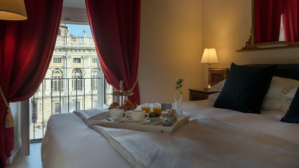 Quarto de hotel com uma cama, um tabuleiro com itens de café da manhã, uma janela com cortinas vermelhas e vista para um prédio lá fora.