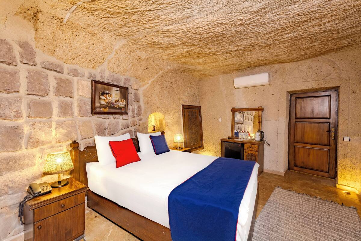 Quarto com parede de pedra, cama de madeira, mesa de cabeceira com abajur e guarda-roupa de madeira.