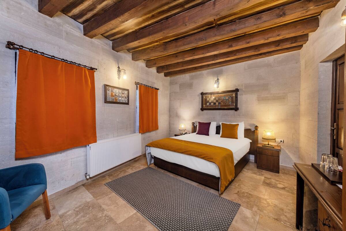 Quarto com cama grande, vigas de madeira, cortinas laranja, mesa de cabeceira com luminária e uma cadeira azul.