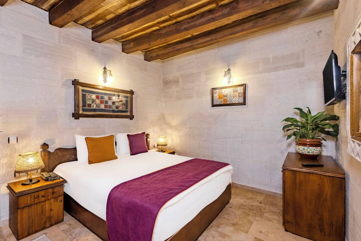 Quarto com teto de madeira, uma cama grande com manta roxa, duas mesas de cabeceira com luminárias e uma planta em vaso.