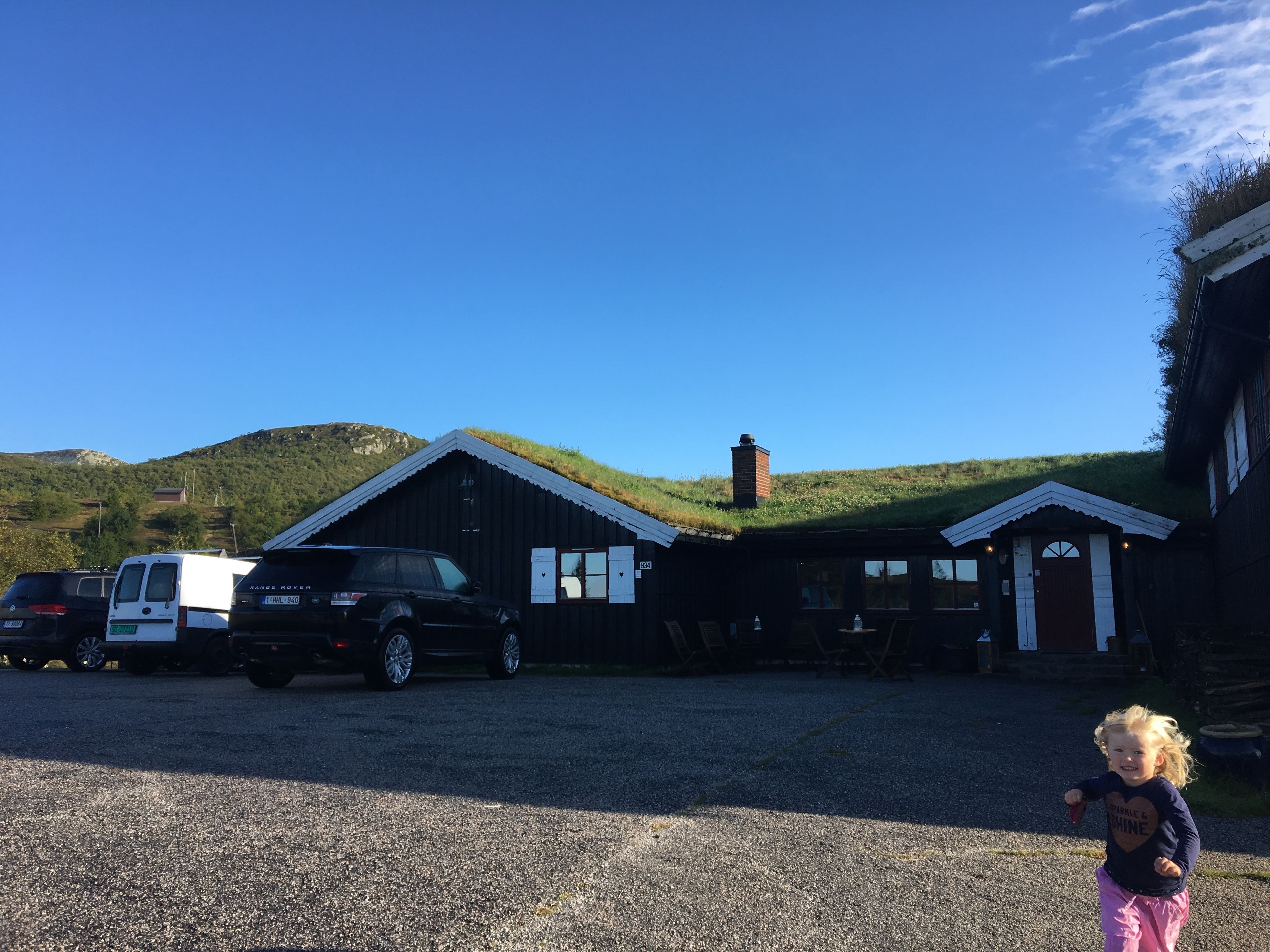 Paisagem rural com um caminho de cascalho, um carro preto e uma van branca estacionados em frente a um edifício com telhado de grama.