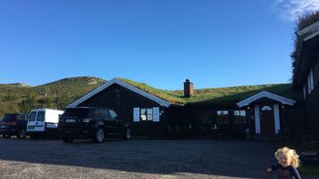 Paisagem rural com um caminho de cascalho, um carro preto e uma van branca estacionados em frente a um edifício com telhado de grama.