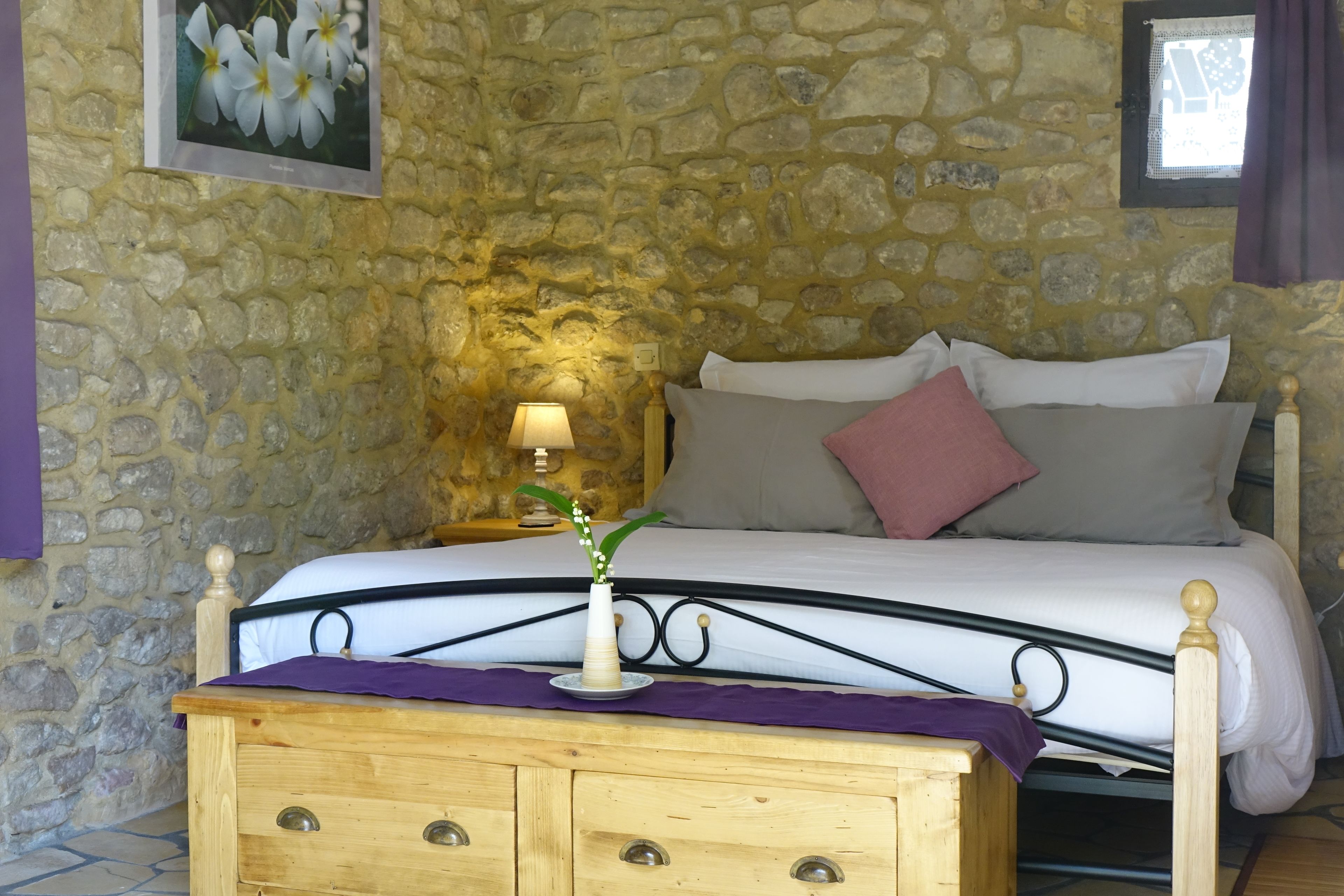 Quarto com parede de pedra, cama com roupa de cama branca, mesa de cabeceira de madeira e luminária.