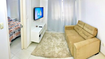 Uma sala de estar com um sofá bege, uma TV sobre um suporte, uma cama estampada e um pequeno armário branco.