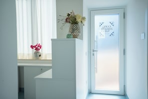 Um quarto com porta branca, janela com cortinas leves, um vaso com flores em uma bancada e uma mesa pequena com um objeto decorativo.