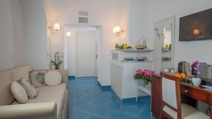 Quarto de hotel com sofá, escrivaninha, cadeira, cafeteira, televisão e uma pequena prateleira com objetos decorativos.