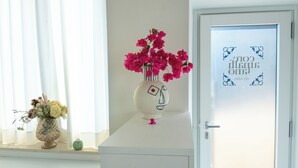 Um quarto com porta branca, janela com cortinas leves e um vaso com flores cor-de-rosa em uma prateleira branca.