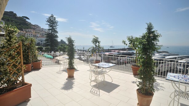 Terraço com vasos de plantas, cadeiras e mesas de metal branco, com vista para uma marina com barcos e uma cidade costeira ao fundo.
