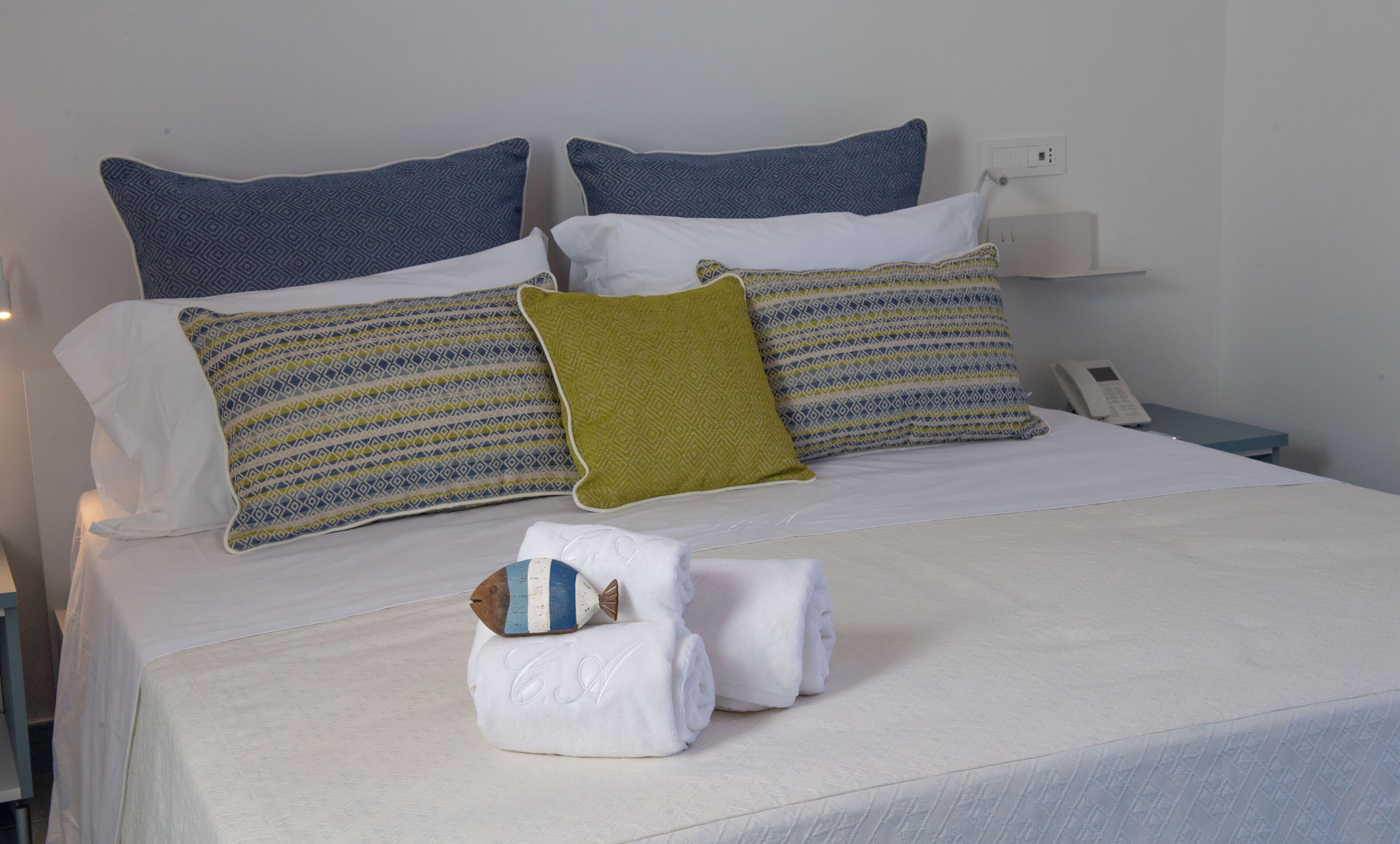 Cama bem arrumada com almofadas estampadas, telefone ao lado e toalhas dobradas sobre um lençol branco.
