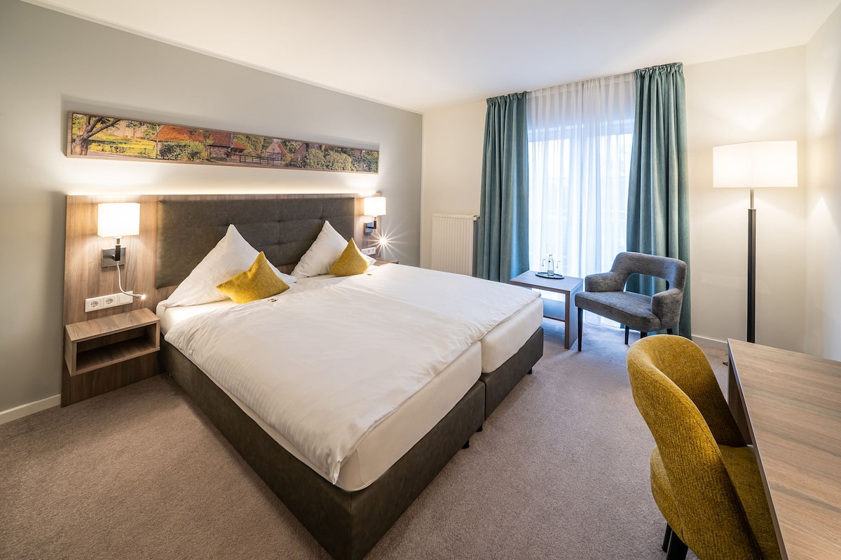 Quarto de hotel com cama grande, luminárias de cabeceira, escrivaninha, cadeira e vista para o exterior.