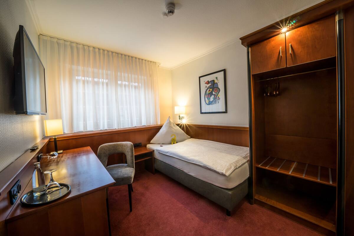 Quarto de hotel com cama, escrivaninha, cadeira, televisão, guarda-roupa e janela com cortinas.