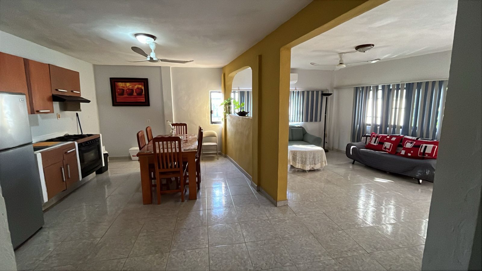 Uma sala de estar ampla com mesa de jantar, cadeiras, sofá e área de cozinha com armários e eletrodomésticos.