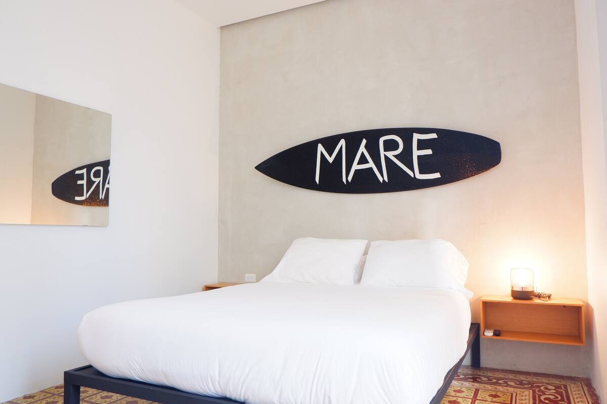 Uma cama bem arrumada com roupa de cama branca, uma mesa de cabeceira com uma luminária e uma placa na parede com a inscrição 'MARE'.