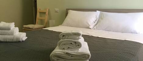 Quarto de hotel com uma cama bem arrumada, uma cadeira de madeira e uma pilha de toalhas dobradas.
