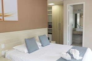 Quarto de hotel com cama, almofadas, mesa de cabeceira, guarda-roupa e banheiro visível por uma porta aberta.