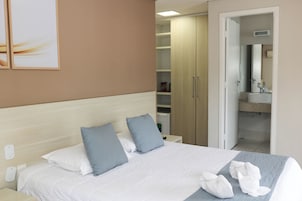 Quarto de hotel com cama, almofadas, mesa de cabeceira, guarda-roupa e banheiro visível por uma porta aberta.