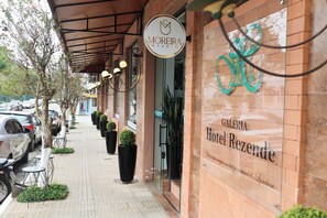 Entrada de hotel com galeria coberta, carros estacionados e plantas em vasos.