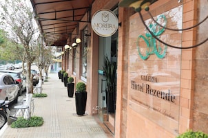 Entrada de hotel com galeria coberta, carros estacionados e plantas em vasos.