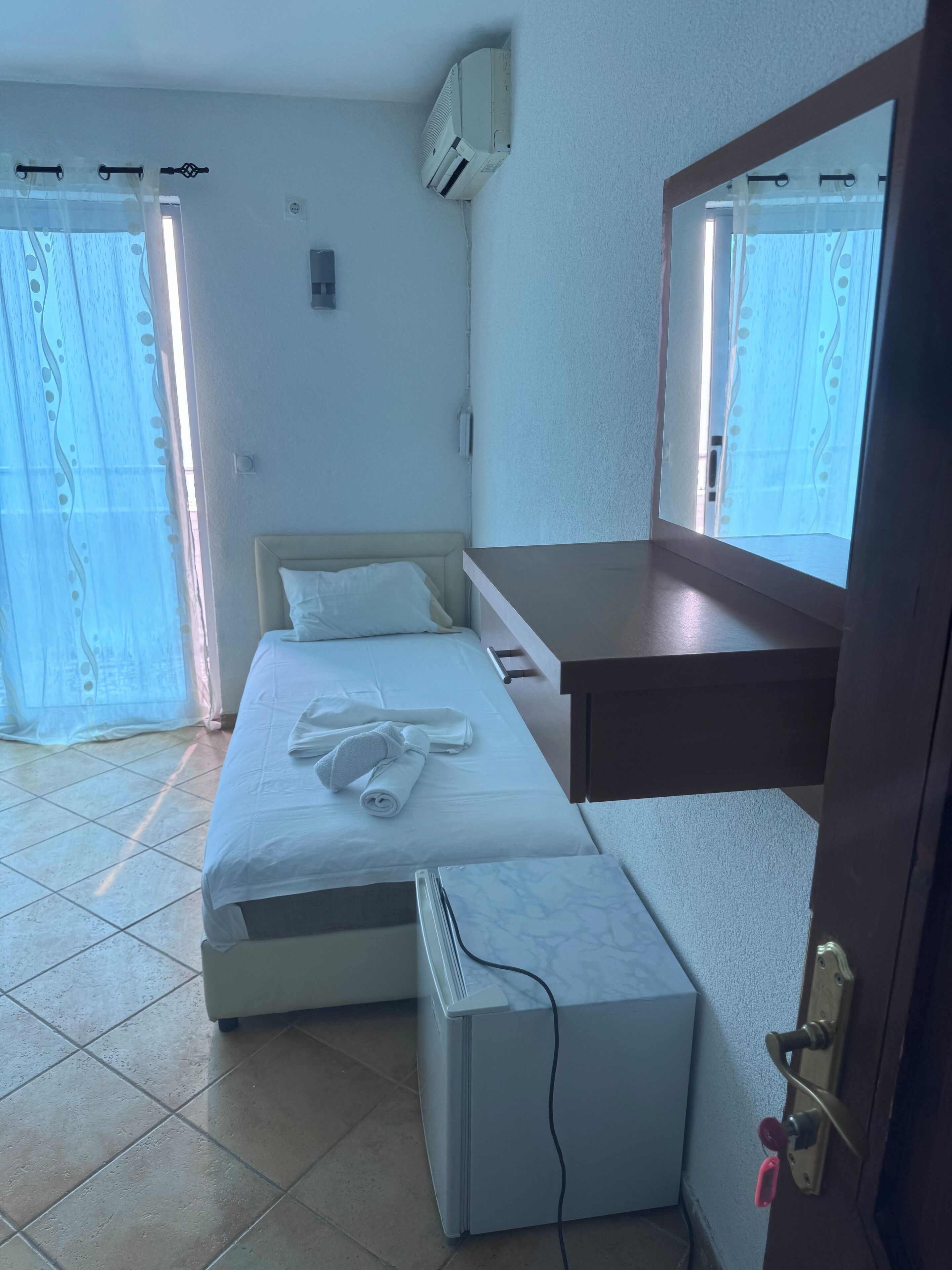 Quarto de hotel com cama, mesa de cabeceira, espelho, ar condicionado e janela com cortinas.