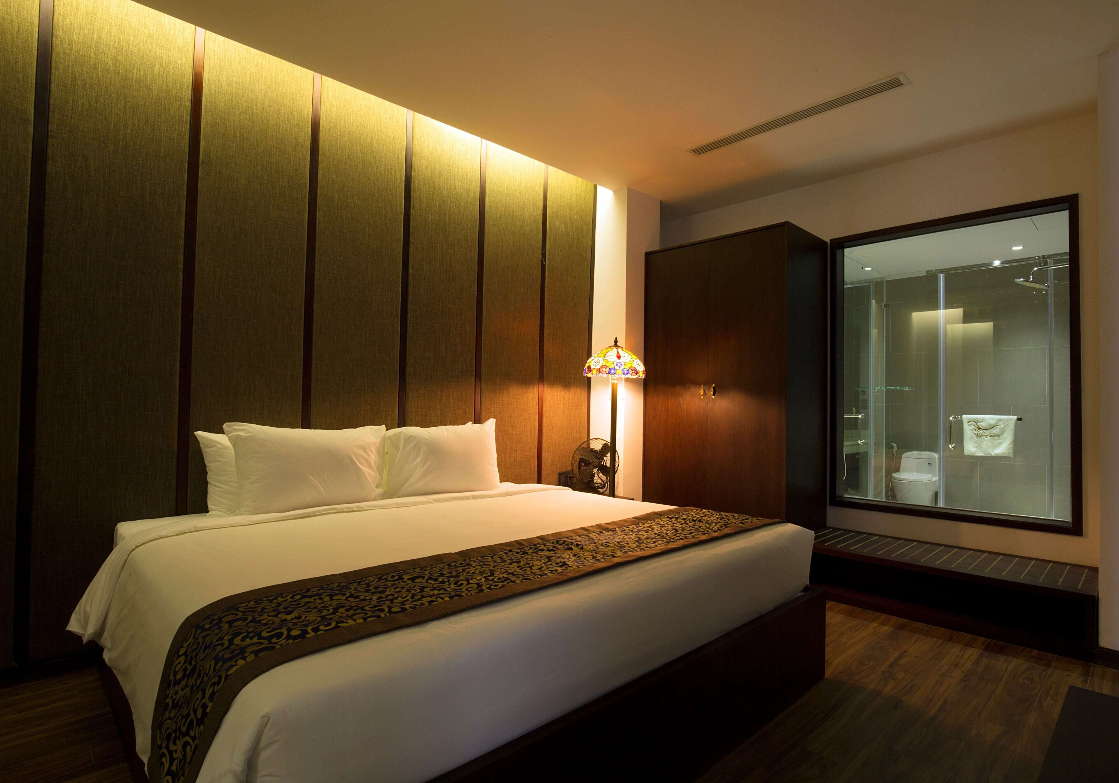 Quarto de hotel com cama grande, mesa de cabeceira, luminária, guarda-roupa e box de vidro.