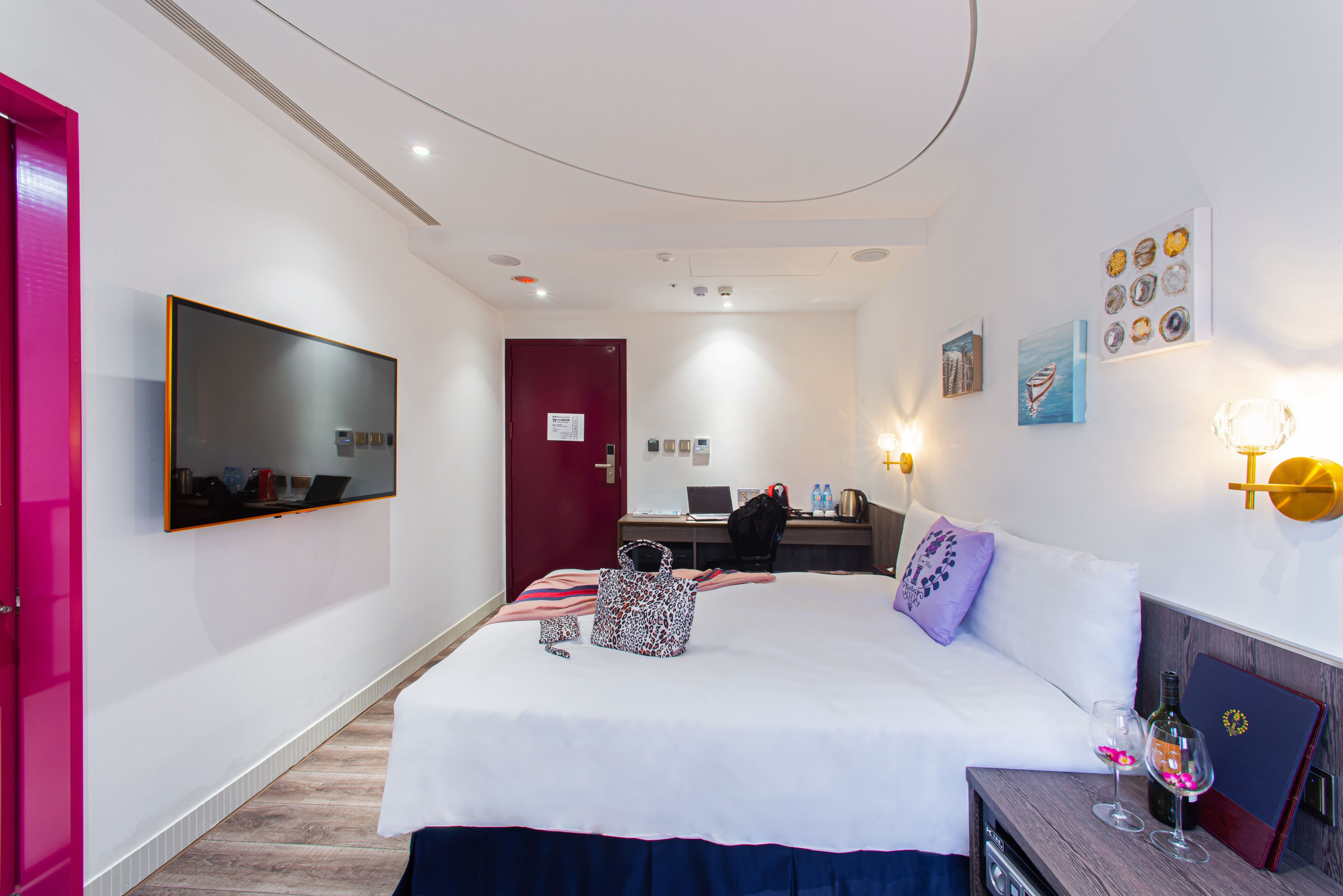 Quarto de hotel com uma cama grande, uma mesa de trabalho com um laptop, uma TV fixada na parede e uma porta com moldura vermelha.