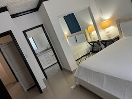 Quarto de hotel com cama, escrivaninha, cadeira, espelho e banheiro com pia e armários.