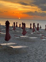 Uma praia com fileiras de espreguiçadeiras e guarda-sóis, um pôr do sol ao fundo, e o oceano.