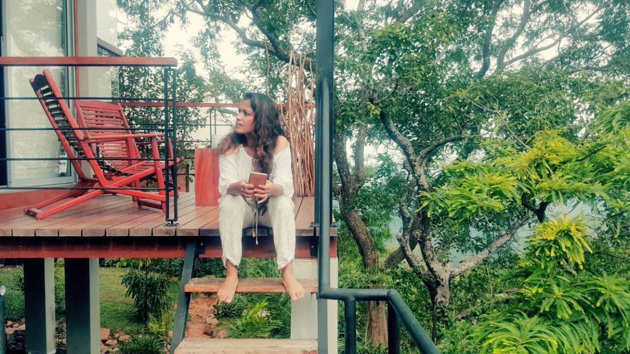 Uma pessoa sentada em um deck de madeira com uma cadeira vermelha, em meio a vegetação exuberante e árvores.