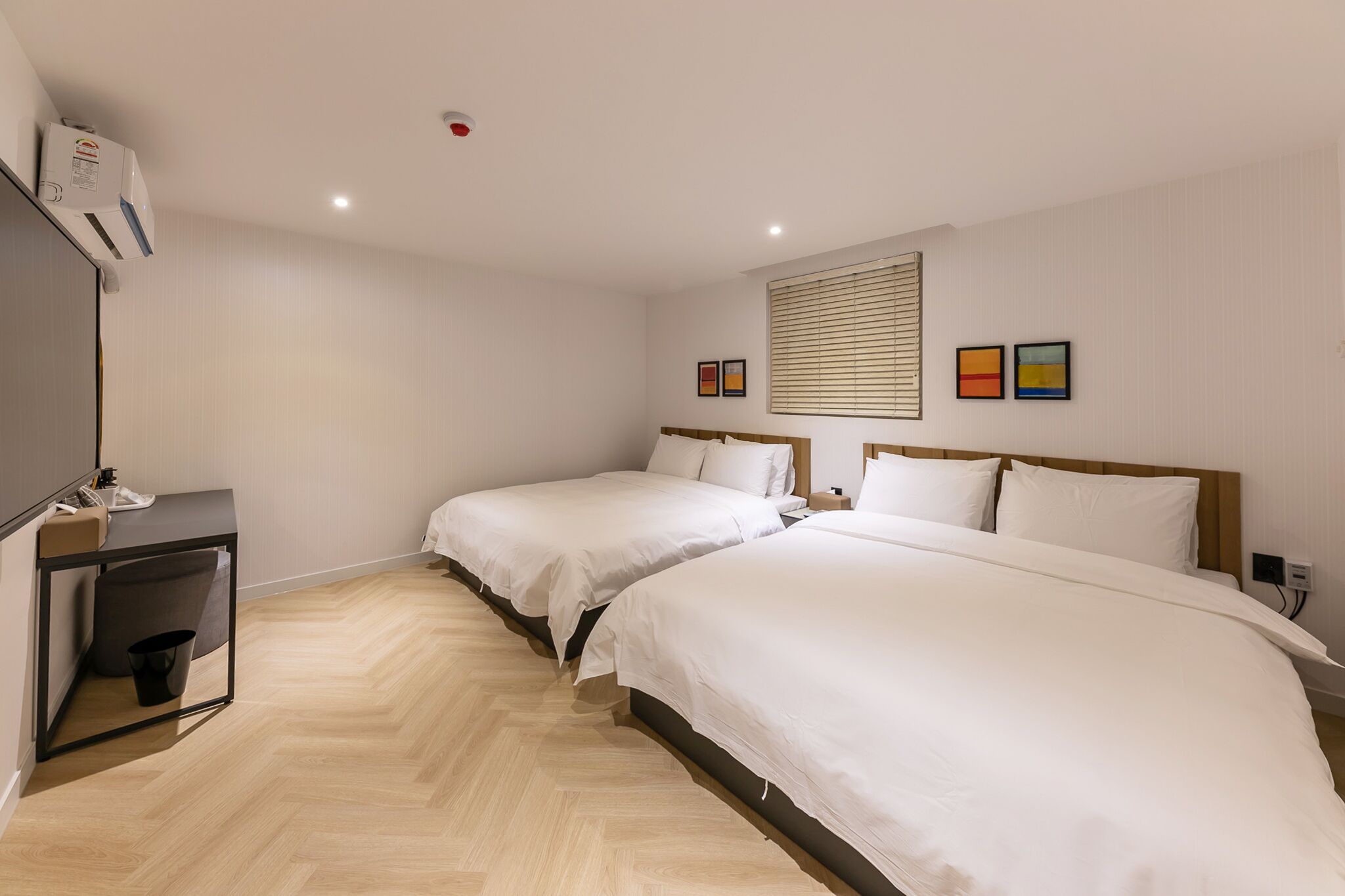 Quarto de hotel com duas camas, uma escrivaninha com cadeira, um cesto de lixo e ar condicionado fixado na parede.