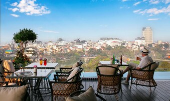 Um terraço no telhado com cadeiras de vime, uma mesa com bebidas e vista para a paisagem urbana.